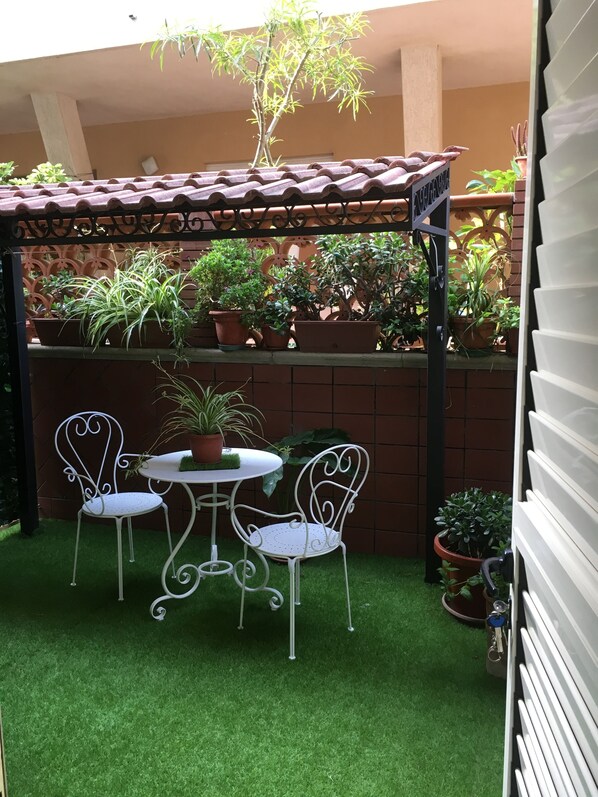 Uma área de pátio com mesa e duas cadeiras, rodeada por plantas em vasos e uma pérgola.