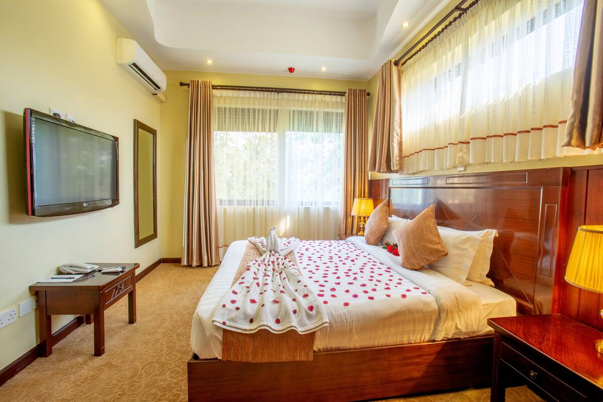 Quarto de hotel com uma cama grande, televisão na parede, escrivaninha com telefone e janela com cortinas.