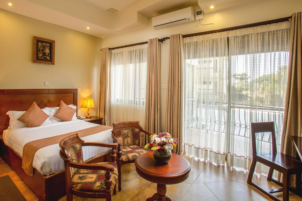 Quarto de hotel com cama, duas cadeiras, mesa com um vaso e varanda com cortinas.