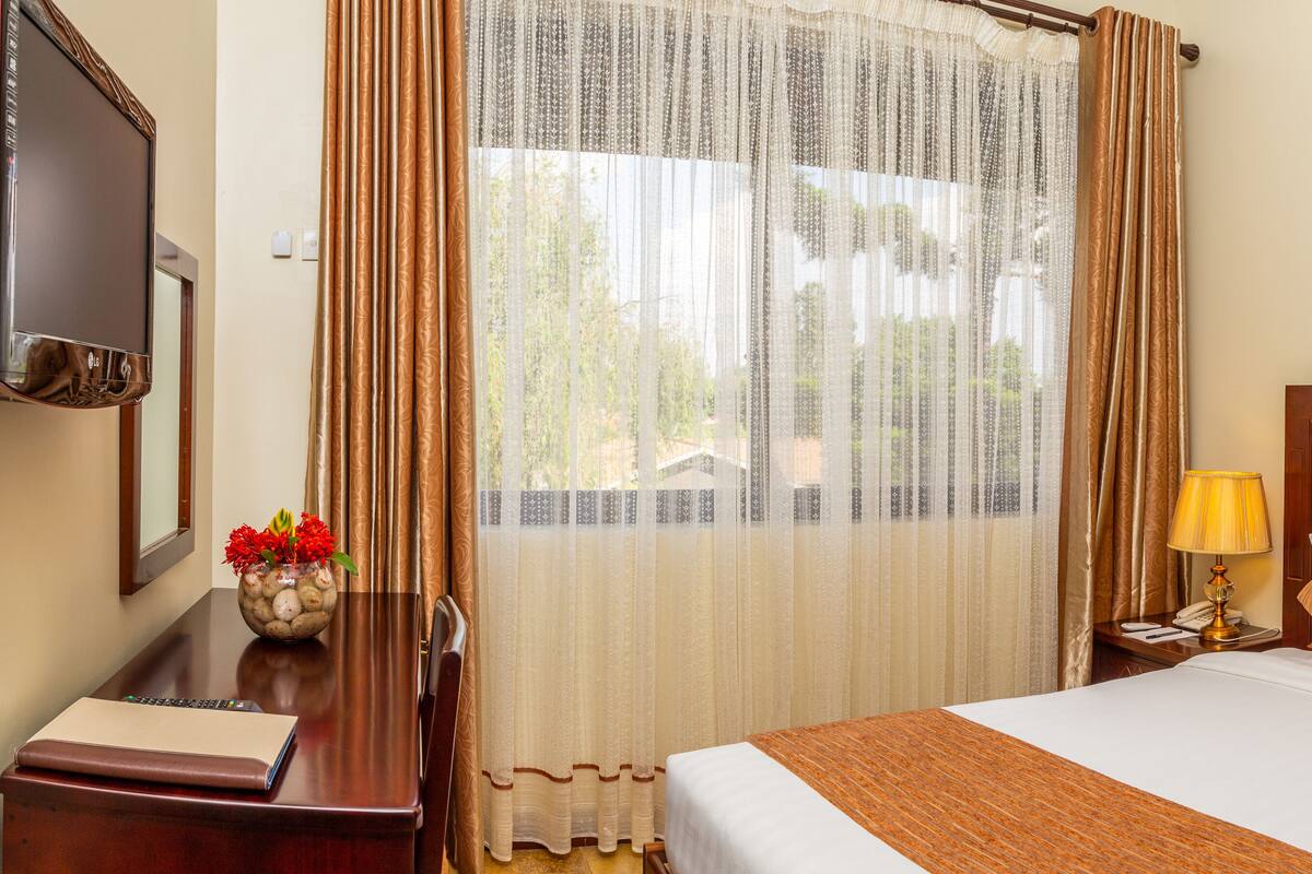 Quarto de hotel com uma cama grande, cabeceira de madeira, duas mesas de cabeceira com abajures, uma TV à esquerda e uma janela com cortinas finas.