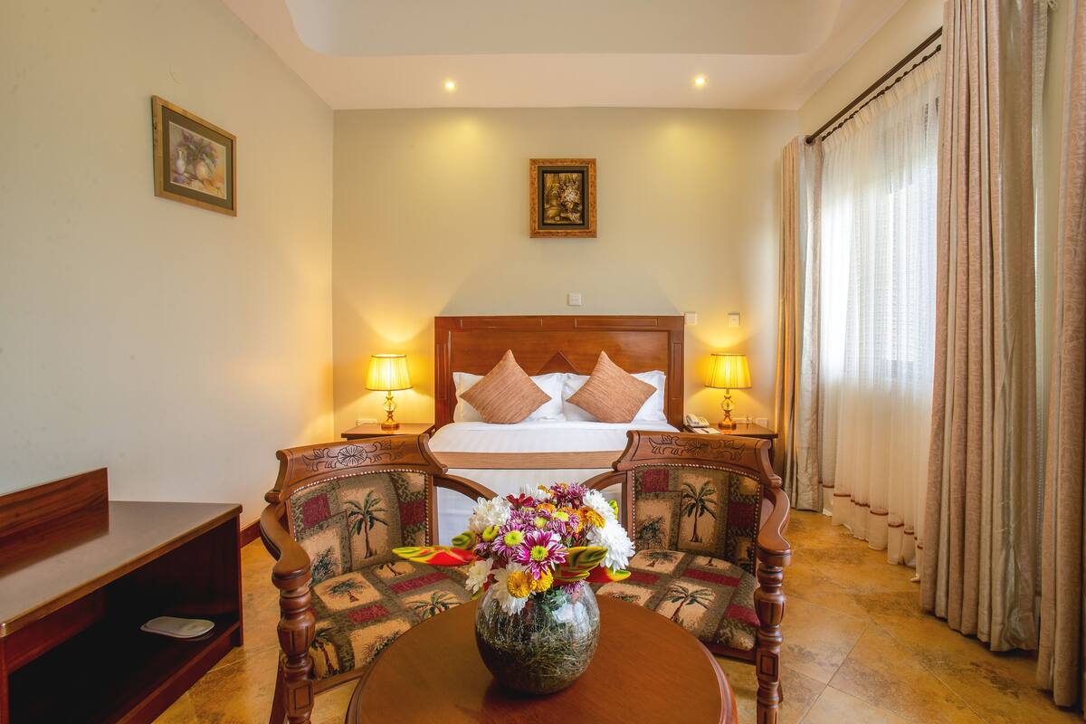Quarto de hotel com uma cama, duas cadeiras, uma mesa com um vaso de flores e uma janela com cortinas.