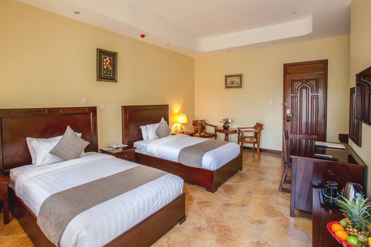 Quarto de hotel com duas camas, uma mesa, uma cadeira, uma mesinha com abajur e uma porta.