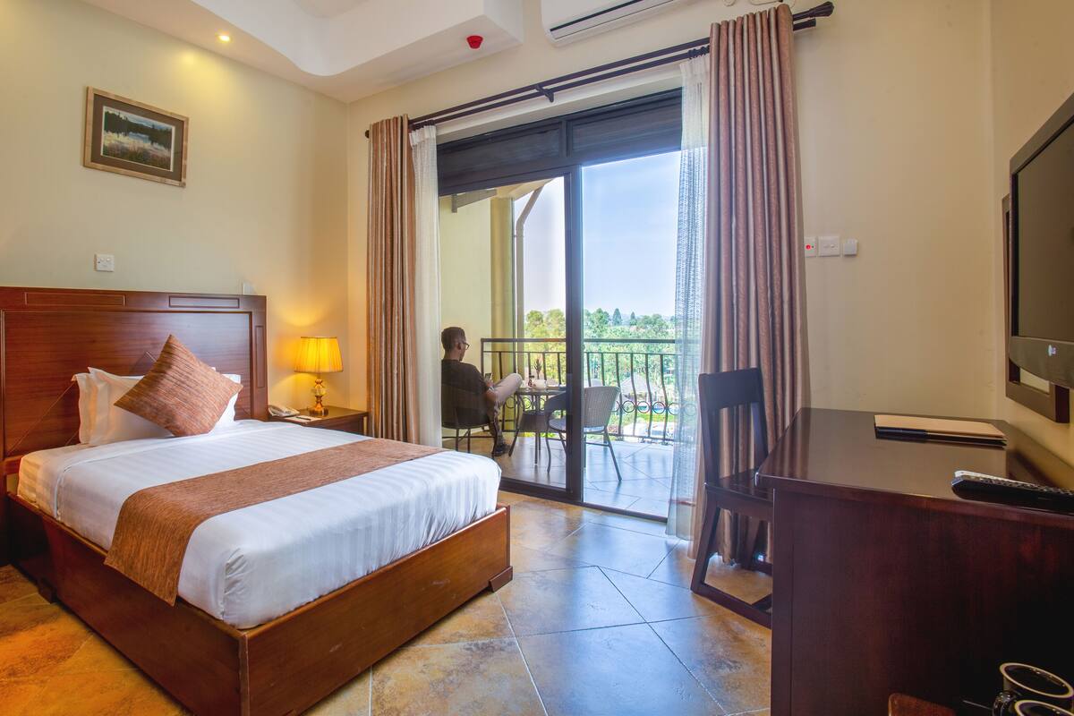 Quarto de hotel com cama, luminárias de cabeceira, escrivaninha com televisão e varanda com vista.
