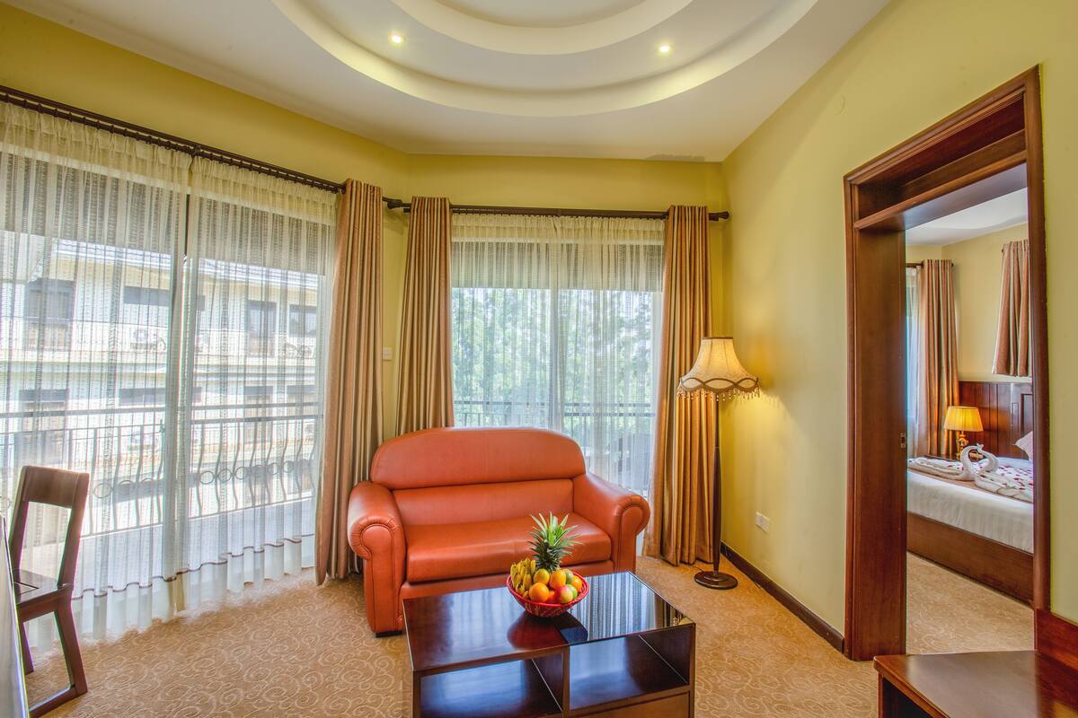 Quarto de hotel com sofá, mesa de centro com frutas, luminária e vista para o exterior através de grandes janelas.