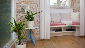 Quarto acolhedor com parede de pedra, sofá branco com almofadas, mesa pequena com uma planta e janela com cortinas.