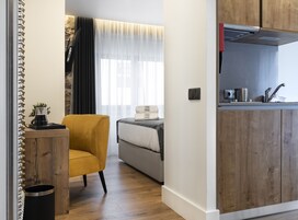Quarto de hotel compacto com cozinha americana, uma cama, um pequeno espaço de estar e janela com cortinas.