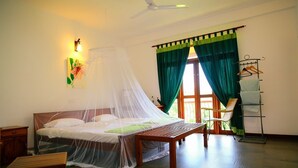 Quarto com cama sob um mosquiteiro, mesa de cabeceira de madeira, uma cadeira, porta-toalhas e uma janela com cortinas verdes.