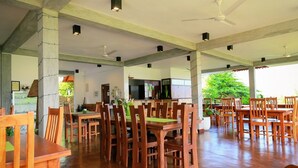 Uma ampla área de refeições com mesas e cadeiras de madeira, ventiladores no teto e uma cozinha ao fundo.