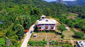 Uma casa de dois andares com telhado plano e uma pequena construção anexa, rodeada por vegetação exuberante e colinas ao fundo.