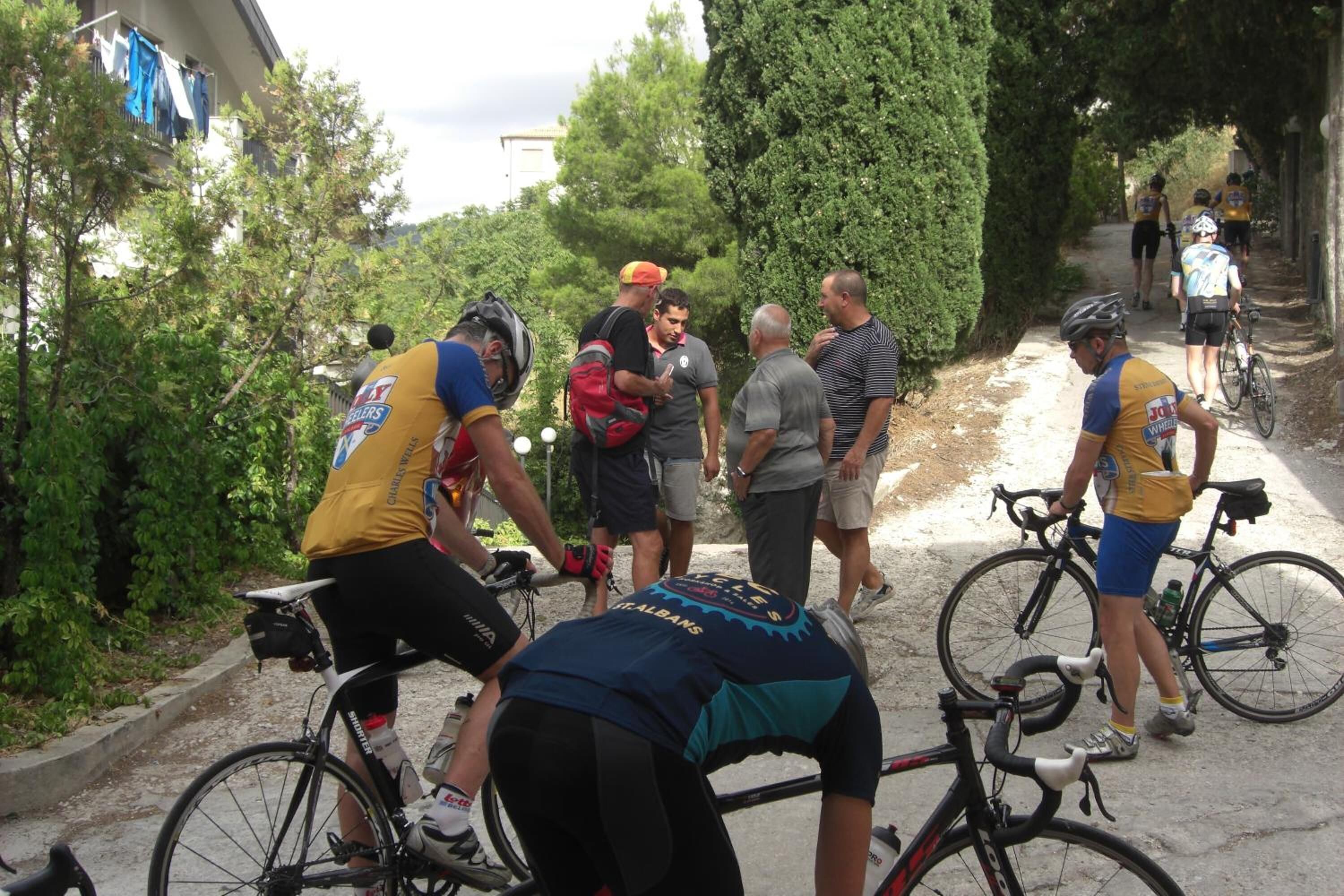 Um grupo de ciclistas reunido numa trilha de terra, com árvores e um edifício ao fundo.