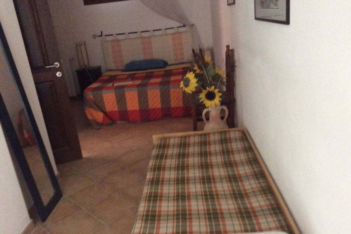 Um quarto pequeno e com pouca luz, com uma cama, uma cadeira e um vaso com girassóis.