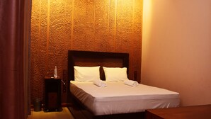 Cama bem arrumada com lençóis e almofadas brancos, mesa de cabeceira com abajur, e parede texturizada ao fundo.