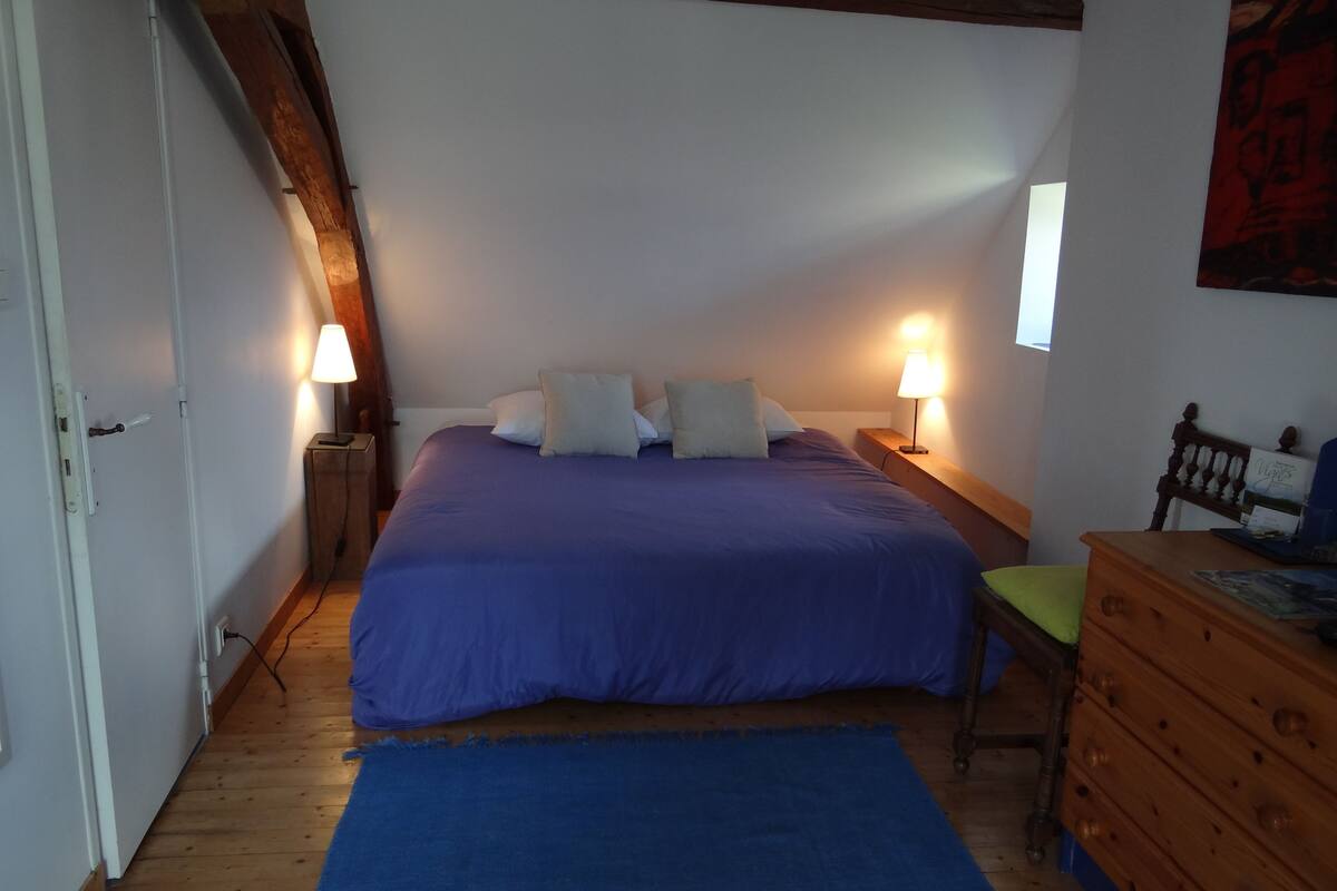 Quarto com cama grande, duas luminárias de cabeceira, cômoda de madeira e tapete azul.