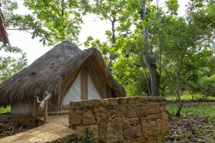 Uma cabana rústica com telhado de palha e parede de pedra à frente, rodeada por vegetação exuberante.