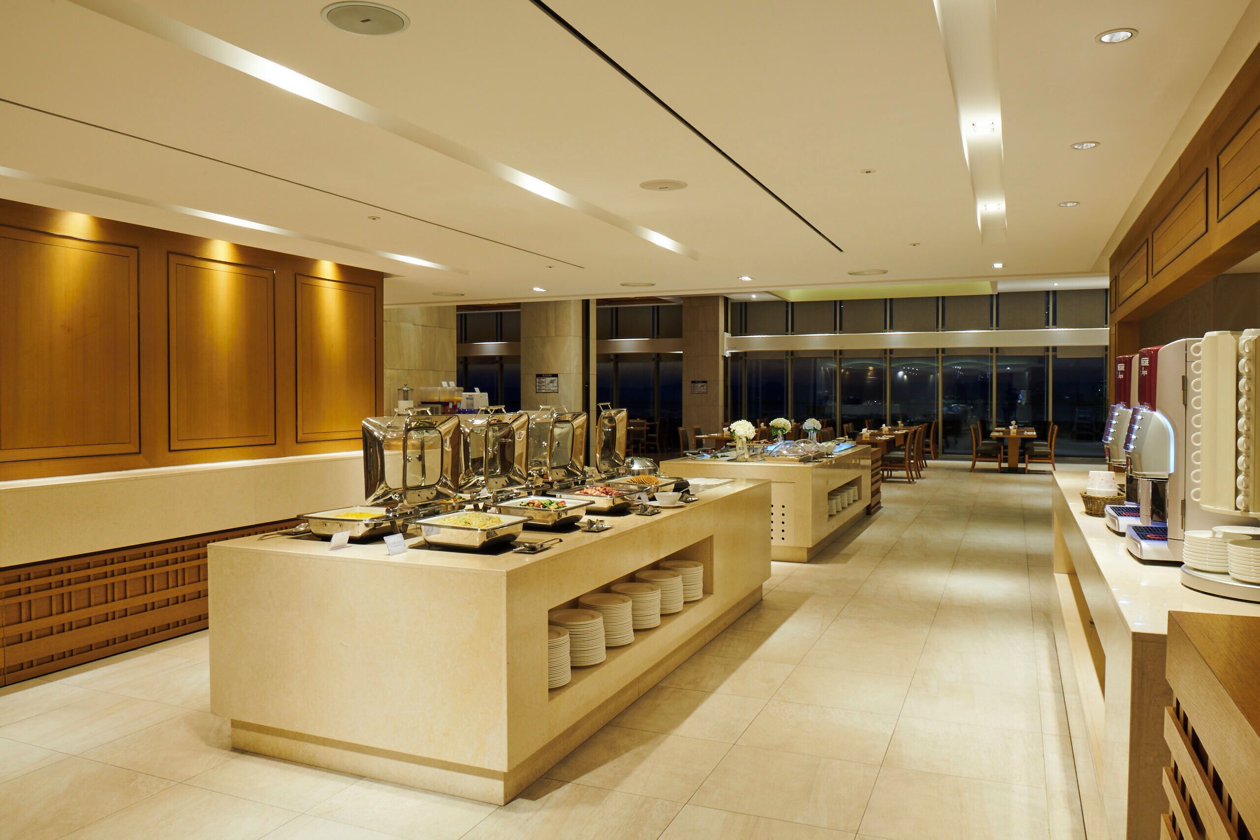 Um lobby de hotel moderno com área de buffet, painéis de madeira e vista para a cidade à noite.