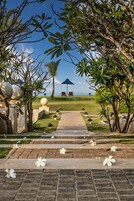 Uma trilha leva a uma área de estar com vista para o mar, cercada por plantas tropicais e um céu limpo.