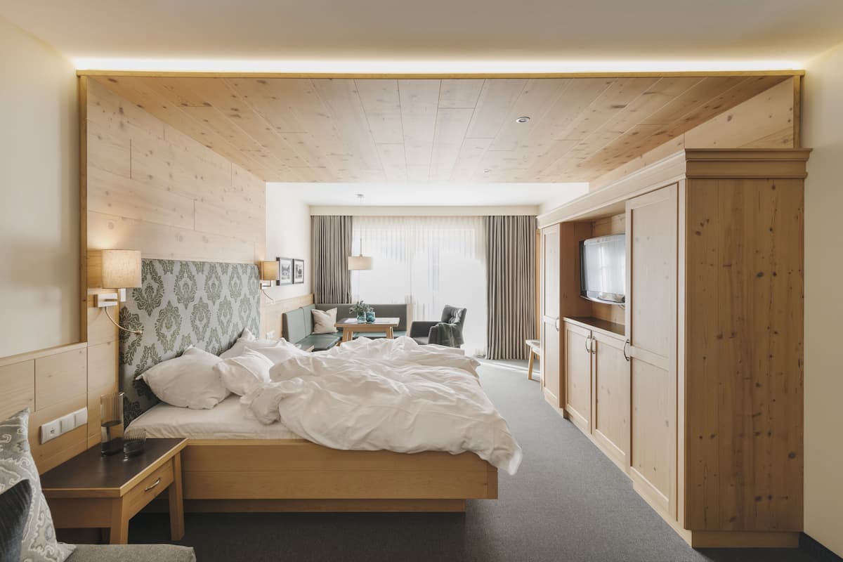 Quarto de hotel com teto de madeira, uma cama grande com roupa de cama branca, uma escrivaninha com abajur, um guarda-roupa e uma televisão.