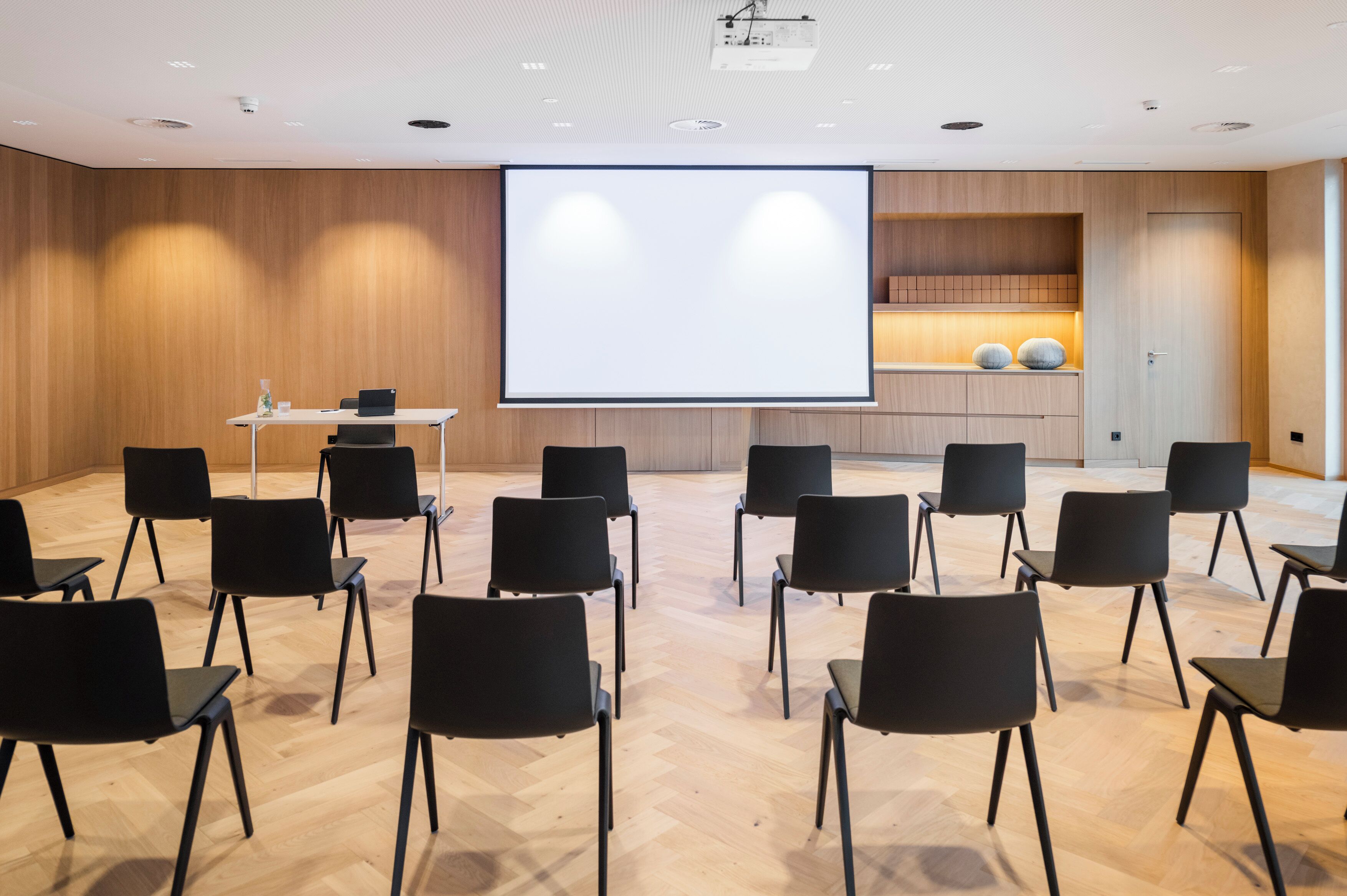 Uma sala de conferências com uma tela de projeção grande, cadeiras pretas dispostas em semicírculo, uma mesa branca com um laptop e um painel de parede de madeira.