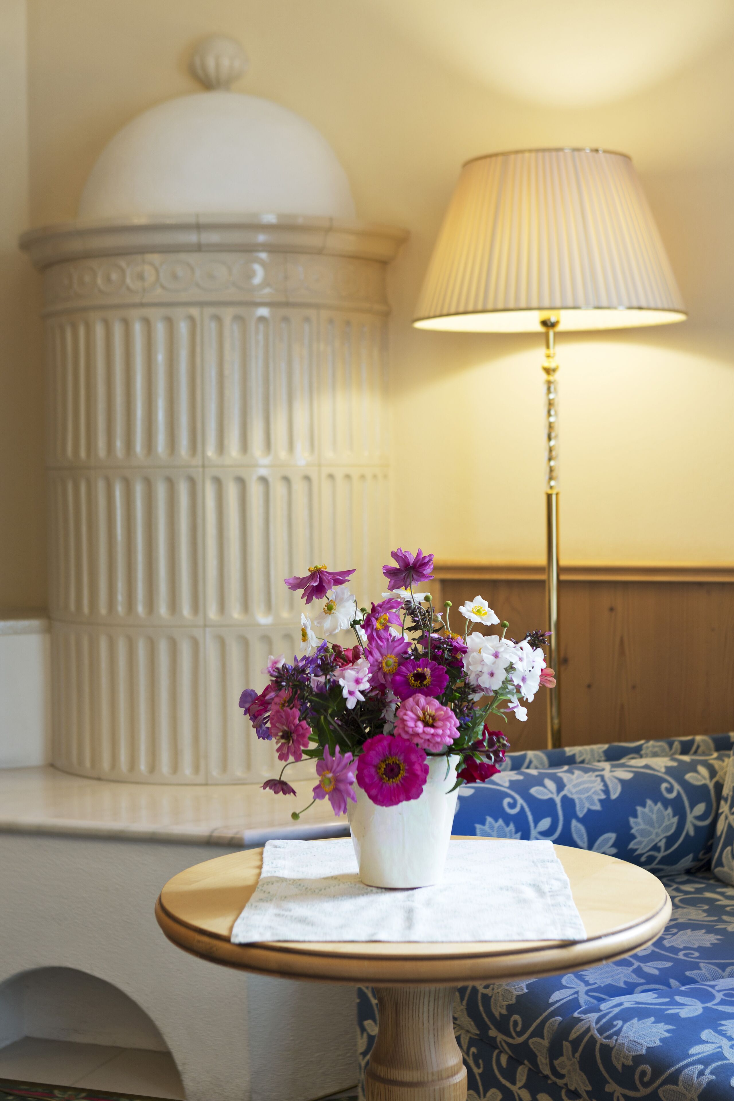 Quarto com uma mesa redonda, um vaso branco com flores, um abajur e um sofá azul estampado.