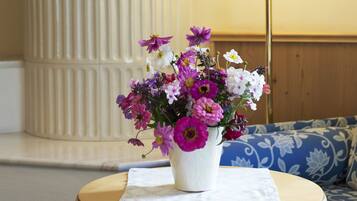 Quarto com uma mesa redonda, um vaso branco com flores, um abajur e um sofá azul estampado.