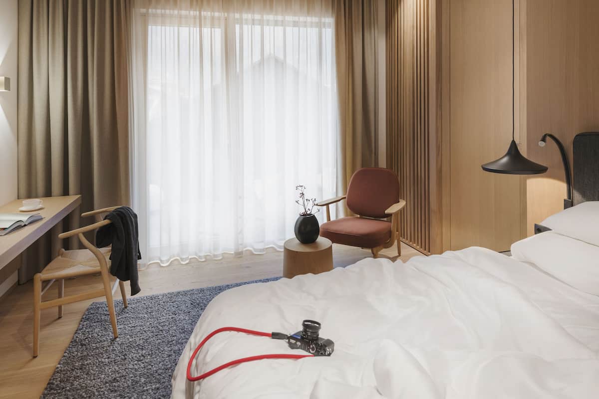 Quarto de hotel com cama, escrivaninha, cadeira, luminária e janela com cortinas.