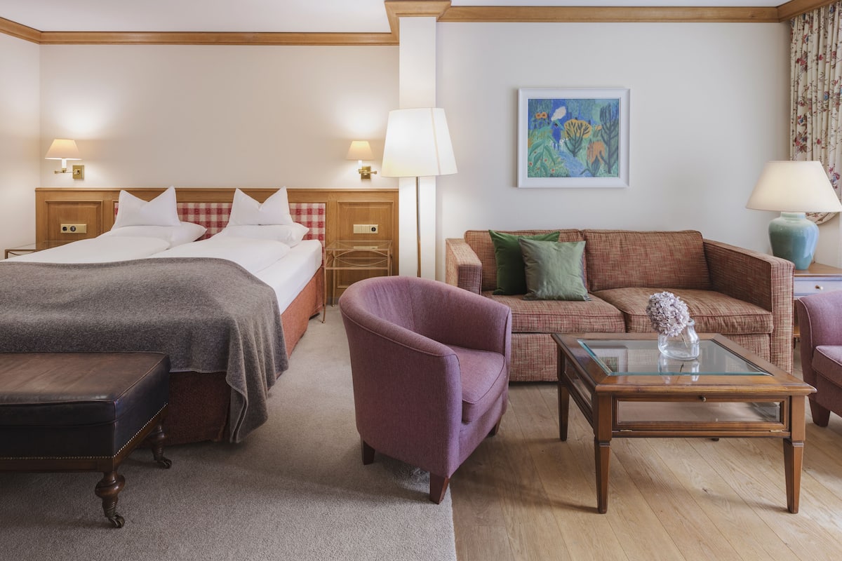 Quarto de hotel com cama, sofá, mesa de centro de vidro e uma cadeira.