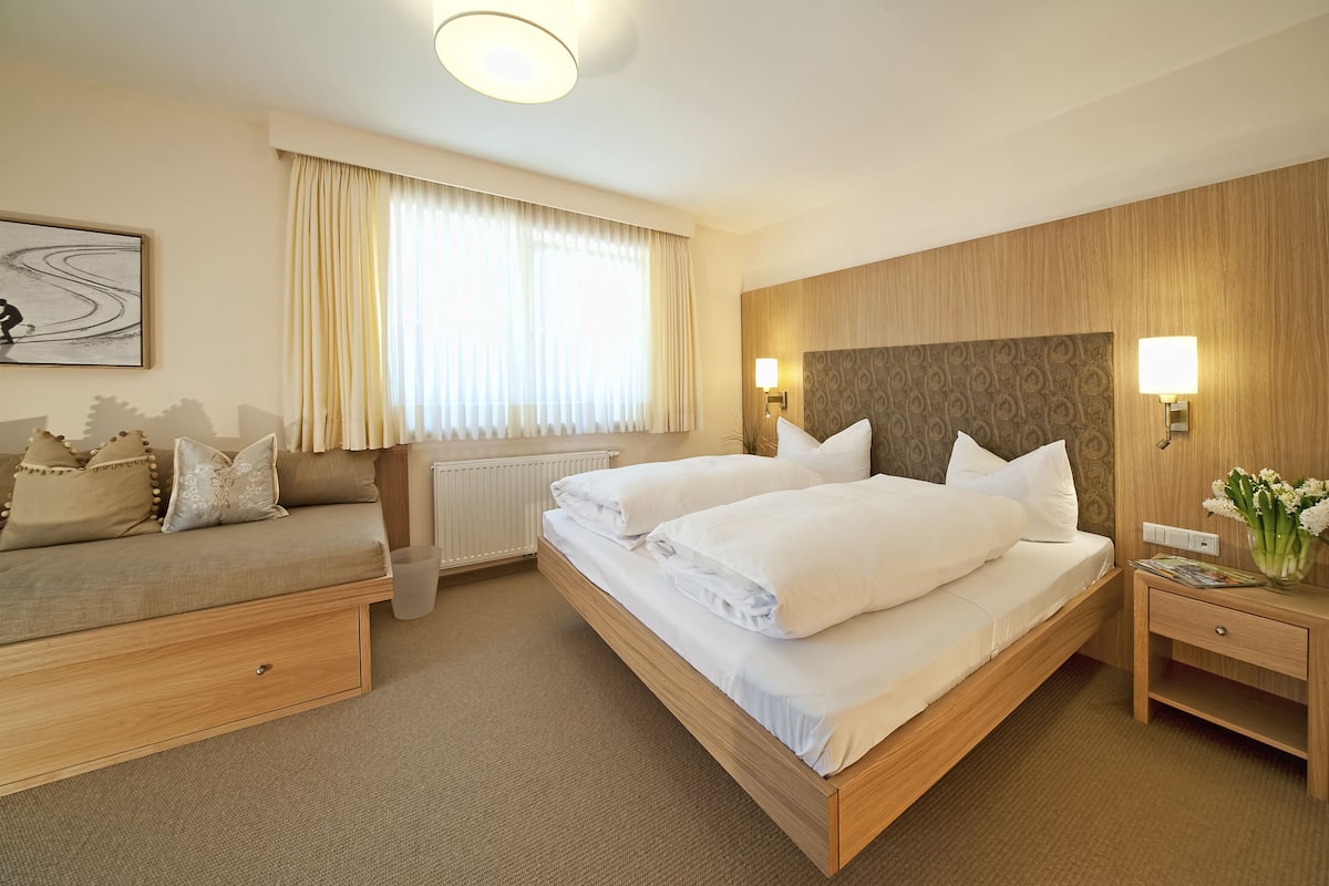 Quarto de hotel com uma cama grande, um sofá, uma mesinha de cabeceira com abajur e uma janela com cortinas.