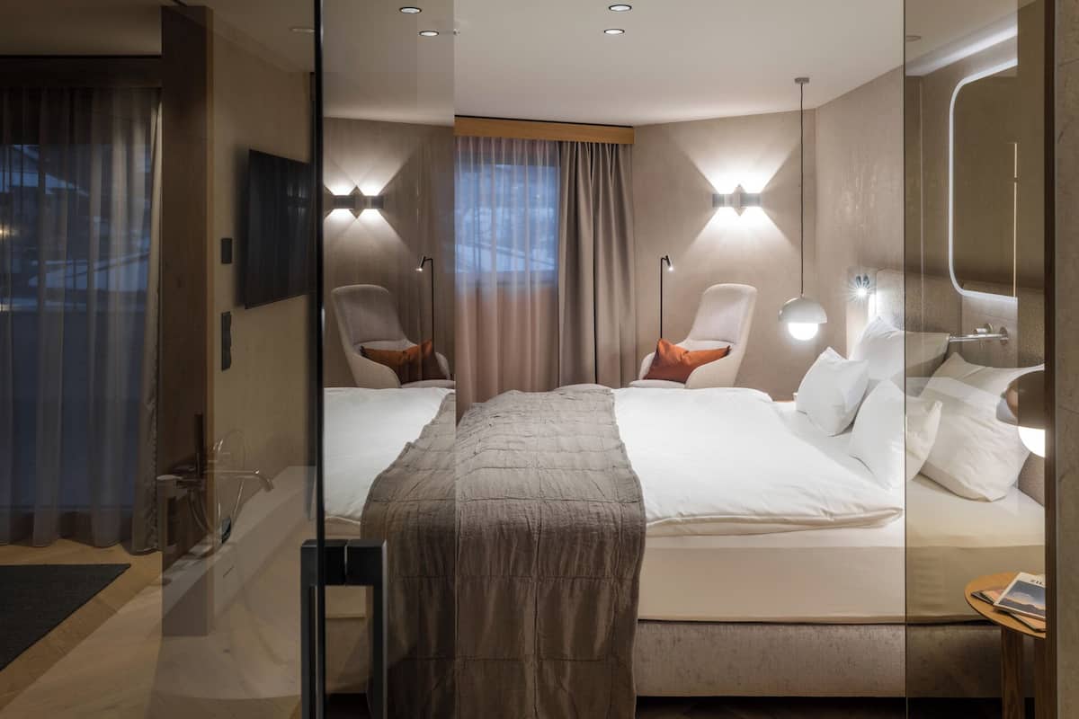 Quarto de hotel moderno com uma cama grande, duas poltronas, uma escrivaninha e um espelho.