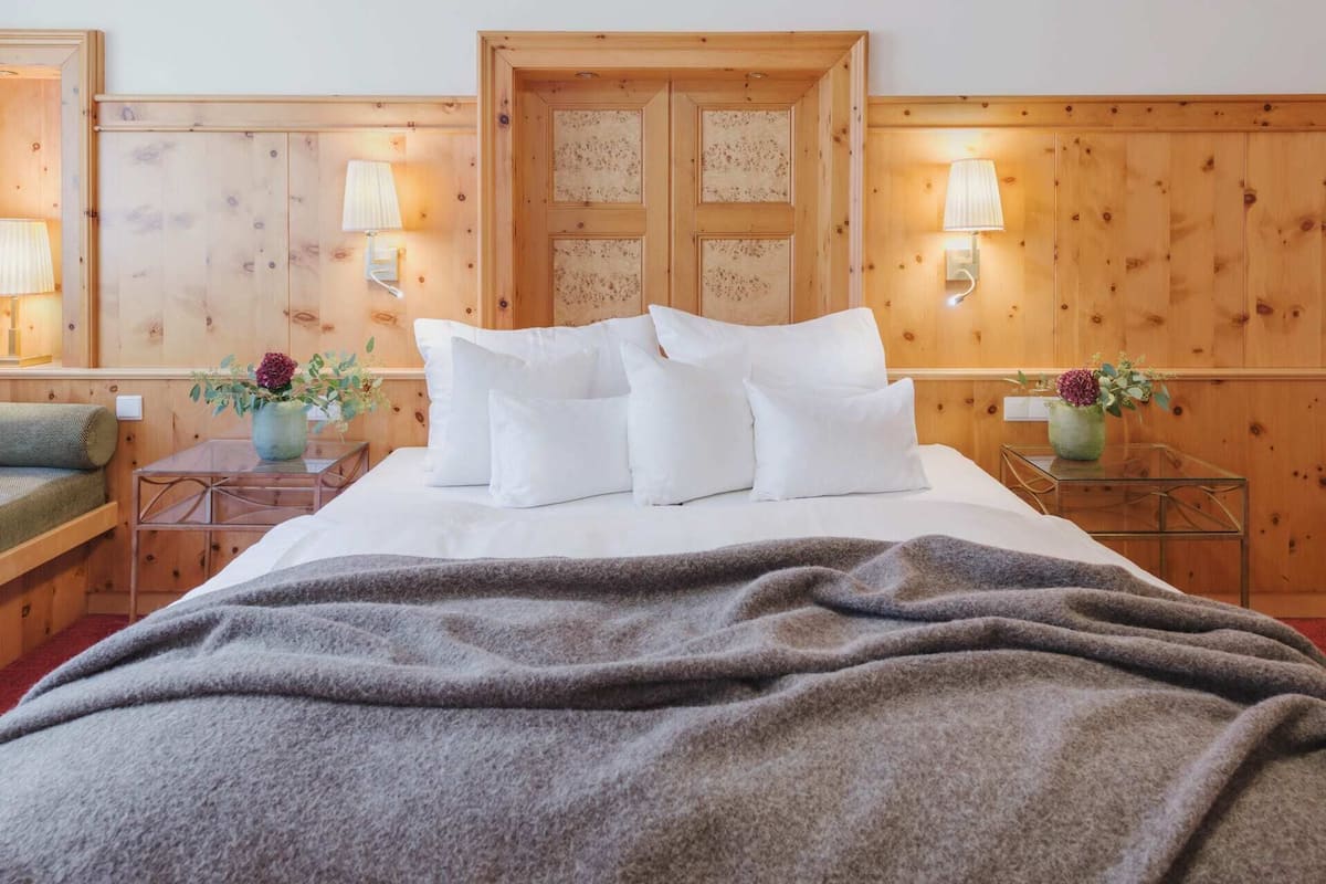 Cama bem arrumada com almofadas brancas e um cobertor cinza, paredes de painel de madeira e duas mesas de cabeceira com luminárias e vasos de flores.