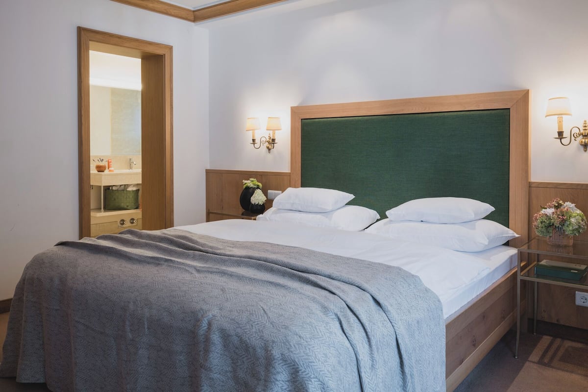 Uma cama bem arrumada com cabeceira verde, duas mesas de cabeceira com luminárias, um espelho e um pequeno vaso com flores.