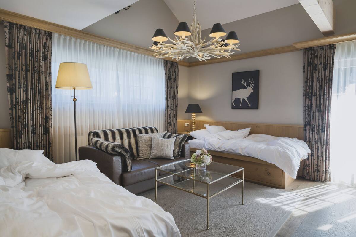 Quarto de hotel com cama, sofá, mesa de centro de vidro e lustre.