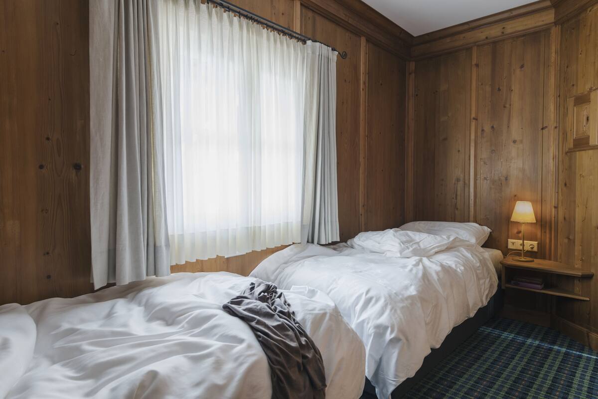 Quarto com duas camas, paredes de madeira, uma janela com cortinas brancas, mesa de cabeceira com abajur e tapete estampado.