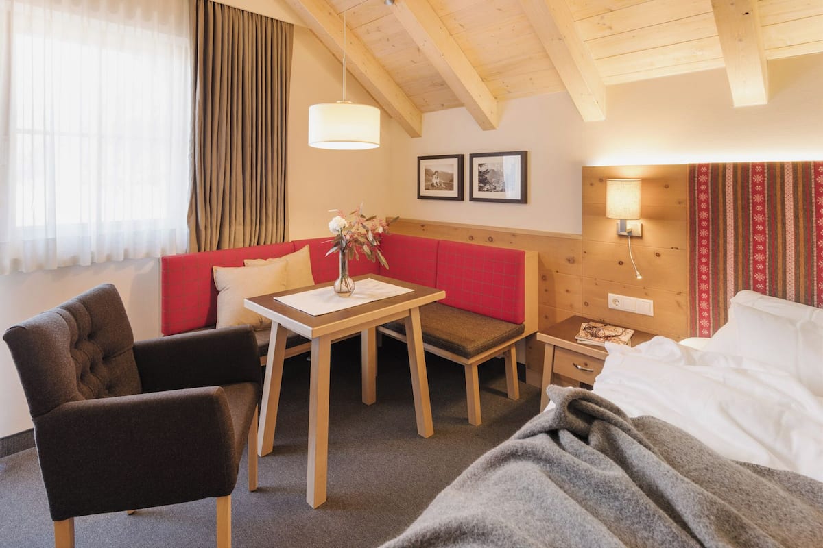 Quarto de hotel com teto de madeira, sofá vermelho, pequena mesa com um vaso de flores, cama com edredom branco e mesa de cabeceira com abajur.