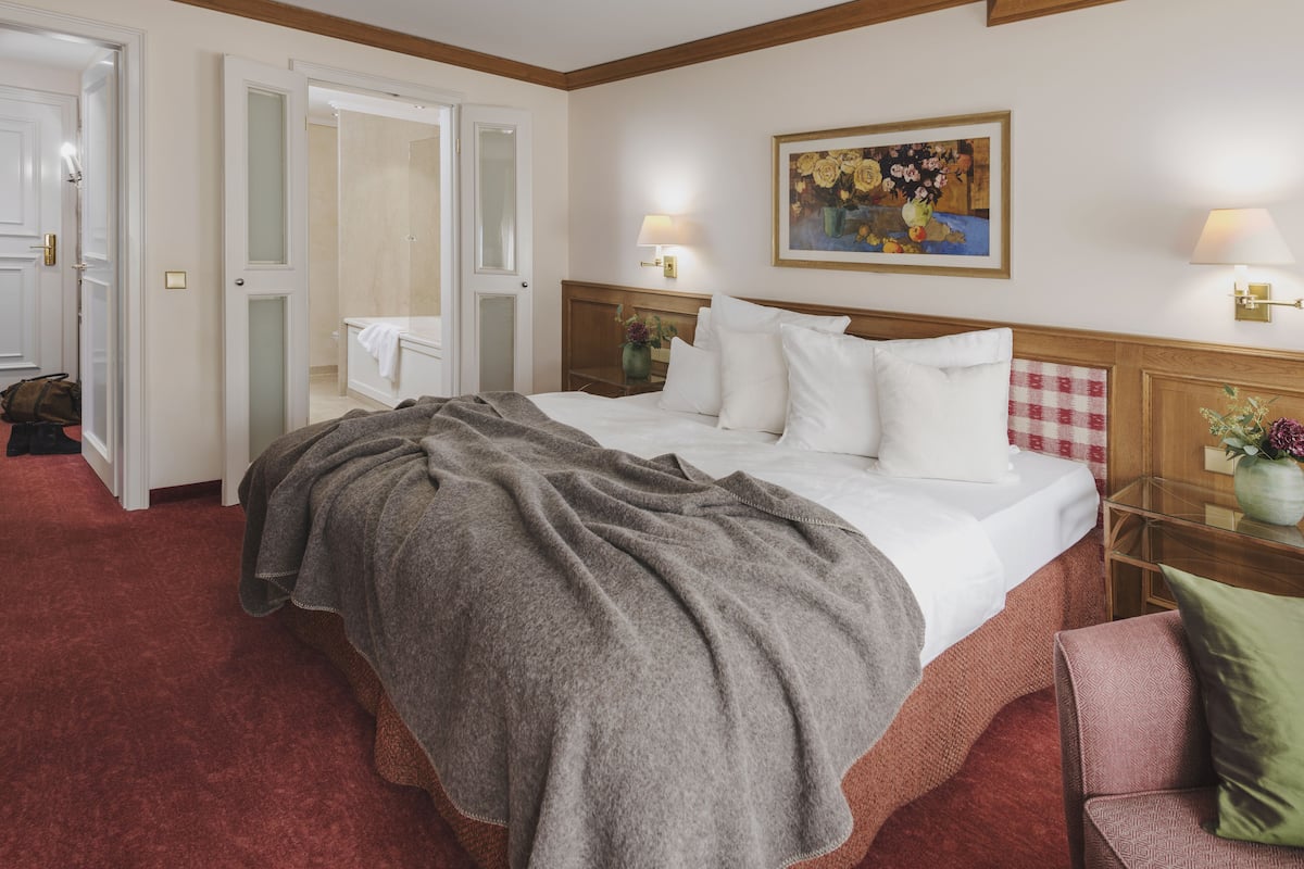 Quarto de hotel com uma cama grande, cabeceira de madeira, duas luminárias laterais, um quadro na parede e um tapete vermelho.