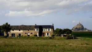 Um edifício de pedra com dois andares, porta vermelha e várias janelas, com um grande castelo no topo de uma colina ao fundo.