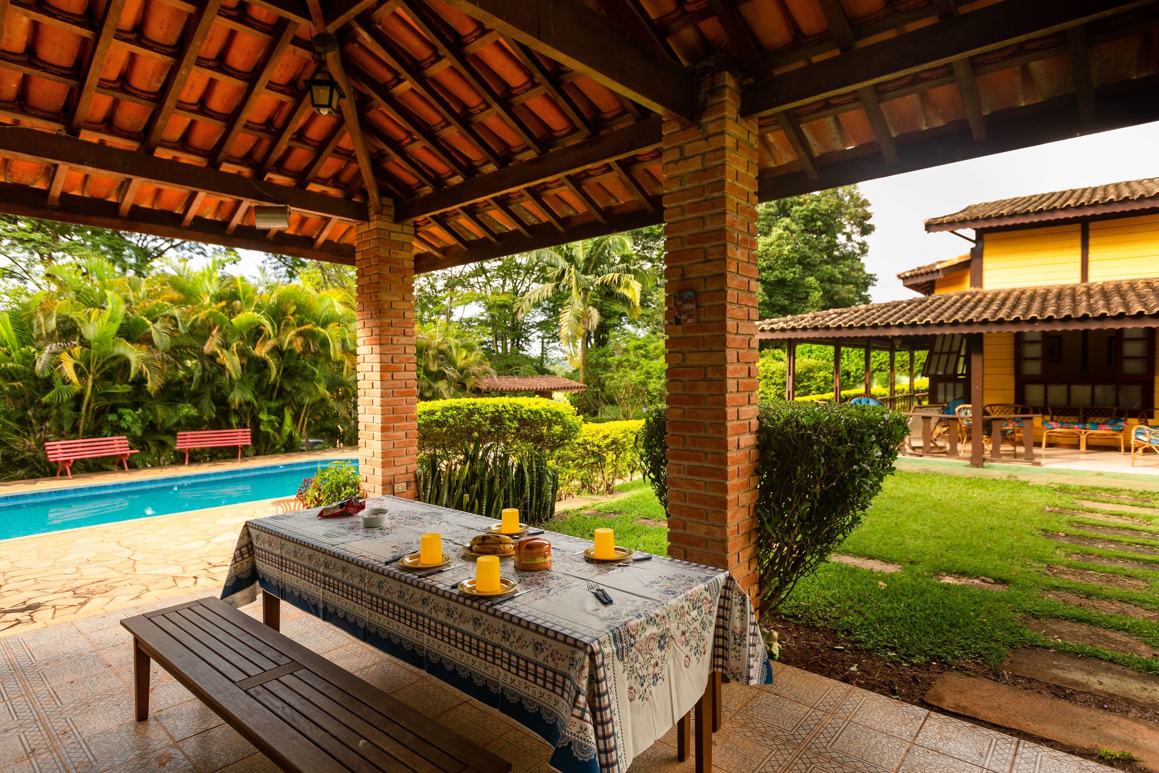 Uma área de refeições ao ar livre coberta, com uma mesa posta para quatro pessoas, rodeada por vegetação exuberante e uma piscina.
