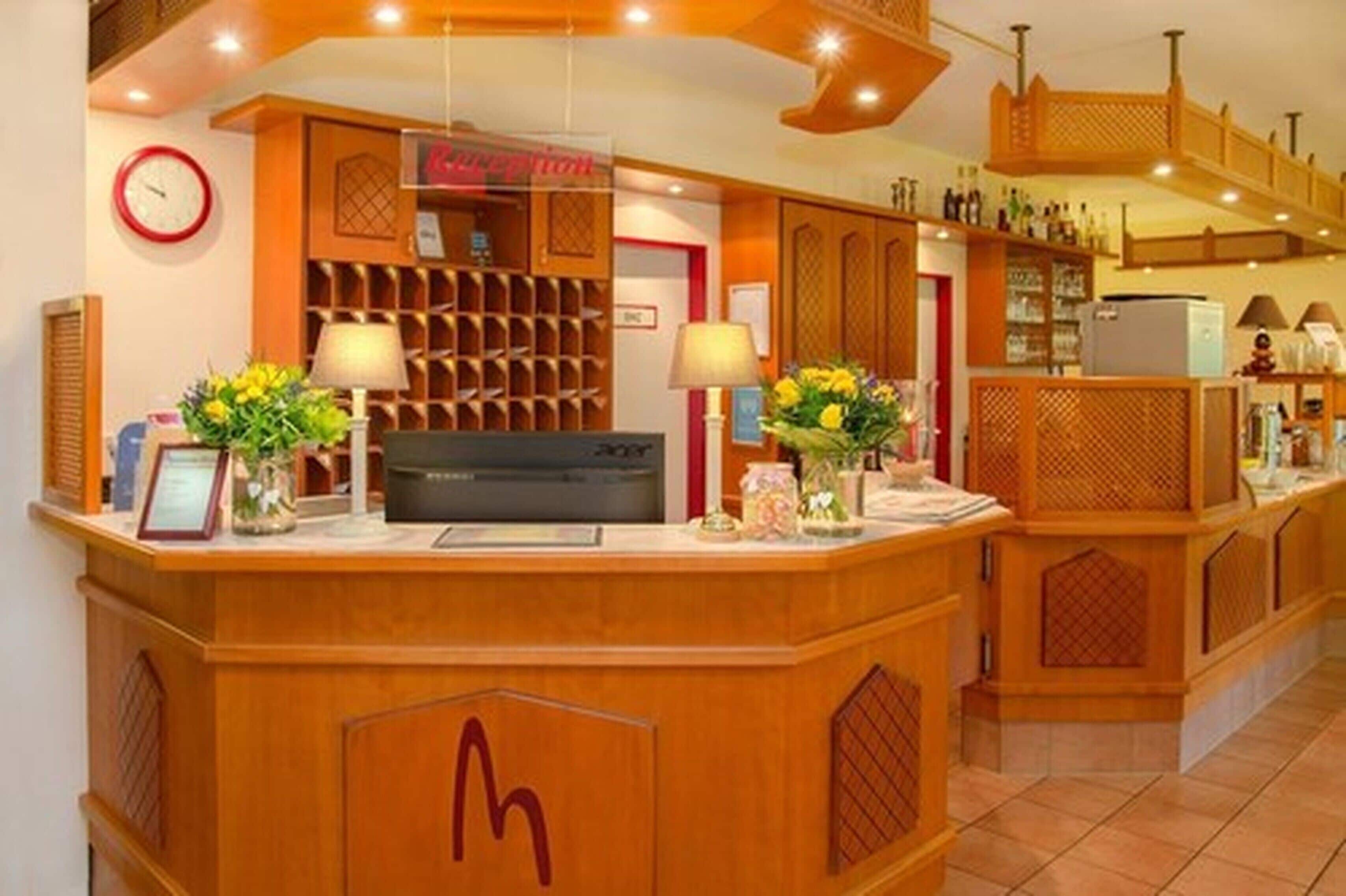 Uma área de recepção de hotel com um balcão de madeira, um relógio na parede e um espaço de bar com prateleiras de garrafas.