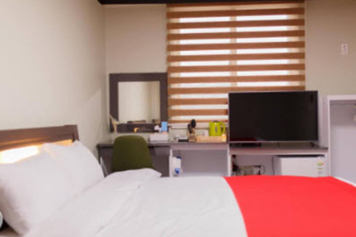 Quarto de hotel com cama, escrivaninha, cadeira, televisão e janela com persianas.