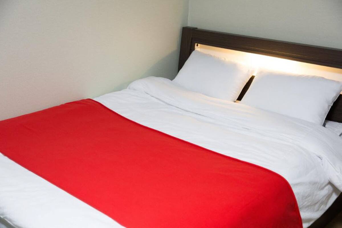 Uma cama com colcha branca e vermelha, cabeceira de madeira e luminária fixada na parede.