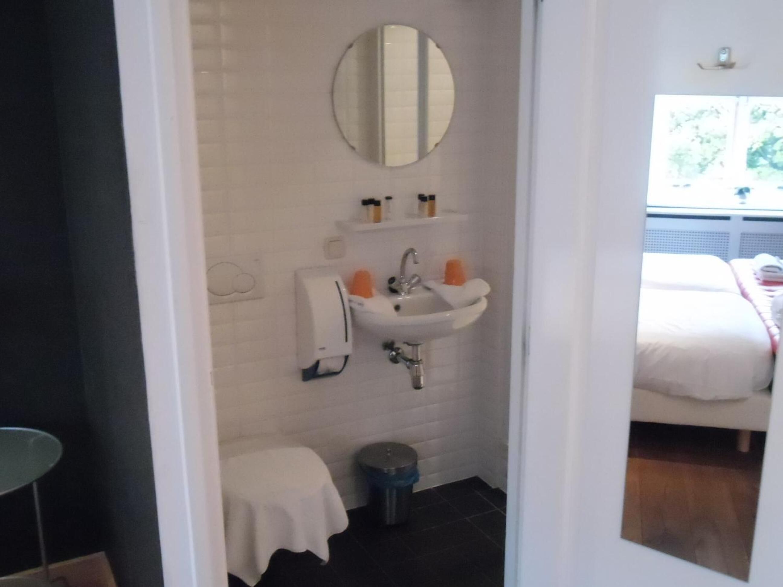 Casa de banho com lavatório branco, espelho e sanita. Há um pequeno contentor de lixo e um porta-toalhas.