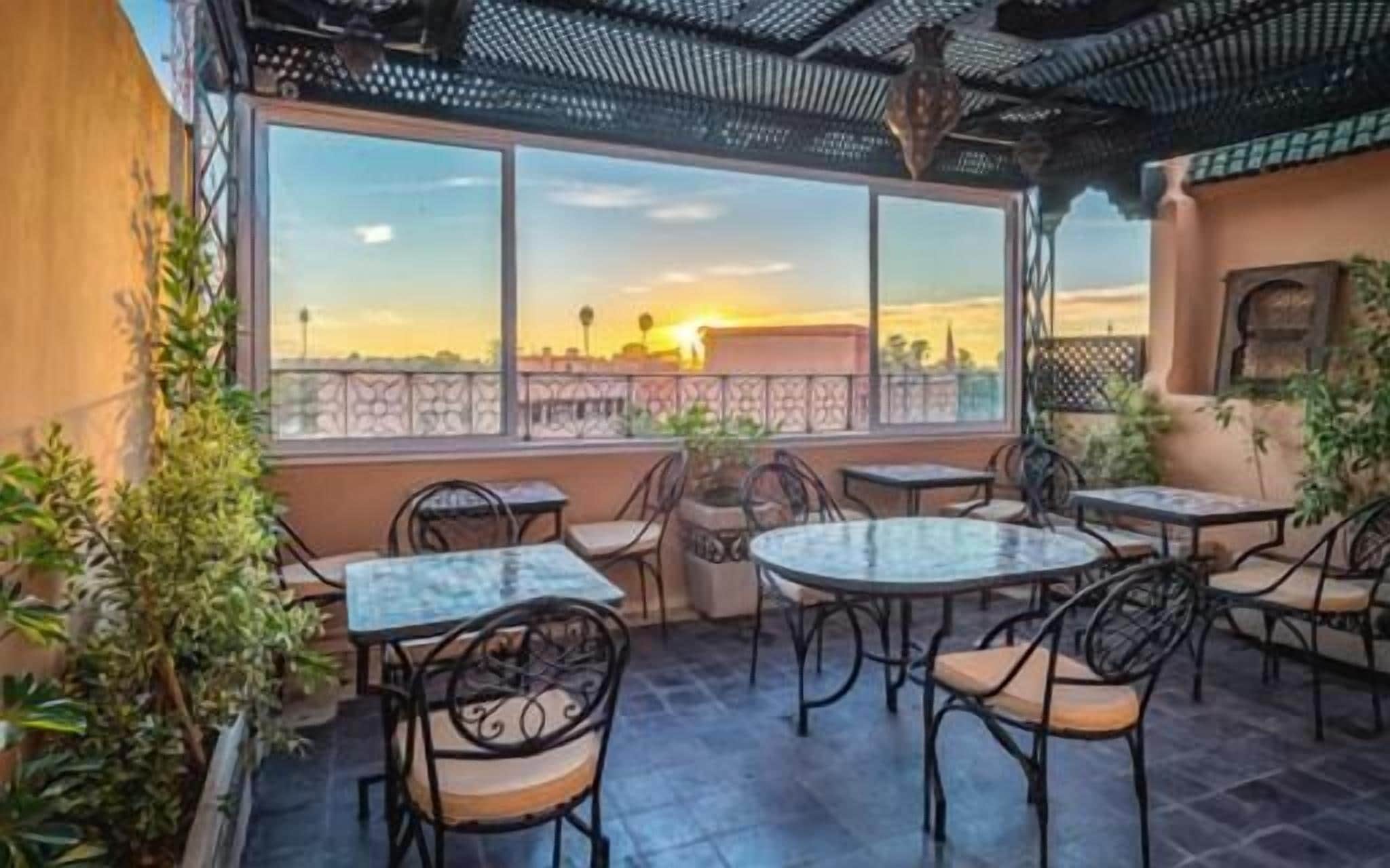 Um terraço no telhado com mesas e cadeiras, grandes janelas com vista para o pôr do sol e plantas decorativas.