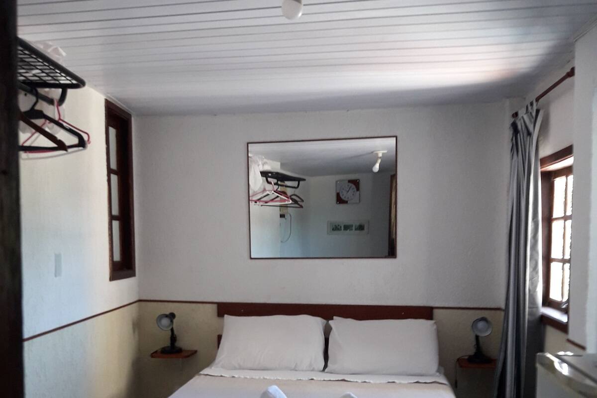 Quarto de hotel com cama, ventilador de teto, espelho, janela com cortinas e mesa de cabeceira com abajur.