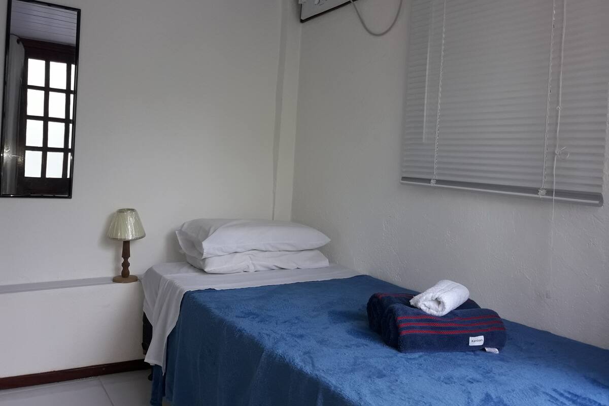 Uma cama de solteiro com colcha azul, um travesseiro branco, uma luminária e uma janela com persianas.