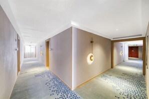Corredor de hotel com carpete estampado, arandelas e uma porta no final.