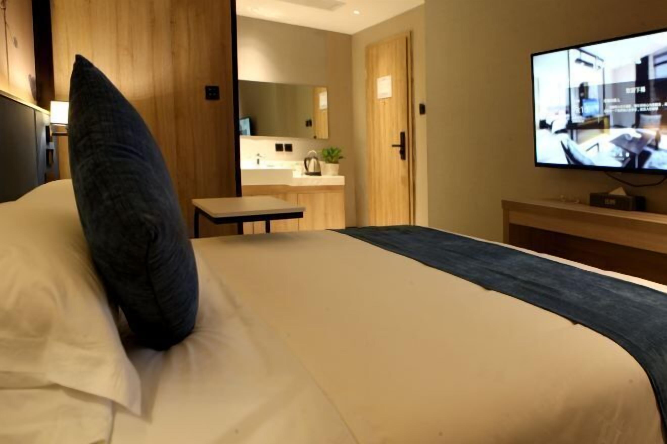 Quarto de hotel moderno com cama, televisão, uma mesinha e banheiro visível ao fundo.