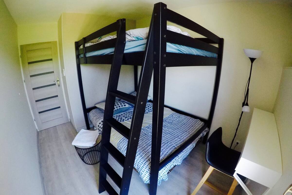 Uma cama beliche com escada, uma escrivaninha com cadeira, uma luminária e uma porta com puxador.