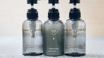 Três garrafas transparentes com bombas dosadoras, identificadas com os números 01, 02 e 03, contendo shampoo, condicionador e sabonete líquido, respectivamente.