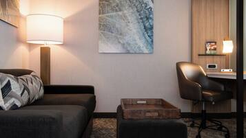 Quarto de hotel com sofá, escrivaninha, cadeira, luminária e um quadro na parede.