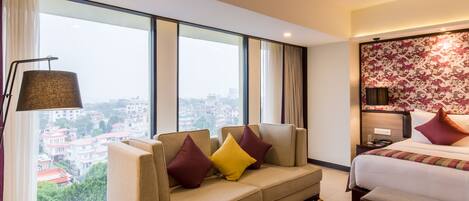 Quarto de hotel moderno com uma janela grande, um sofá com almofadas coloridas, uma cama com cabeceira floral e uma mesa de cabeceira com abajur.