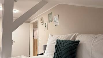 Um quarto com uma cama, um travesseiro, uma mesinha de cabeceira e uma porta que dá para outro cômodo.
